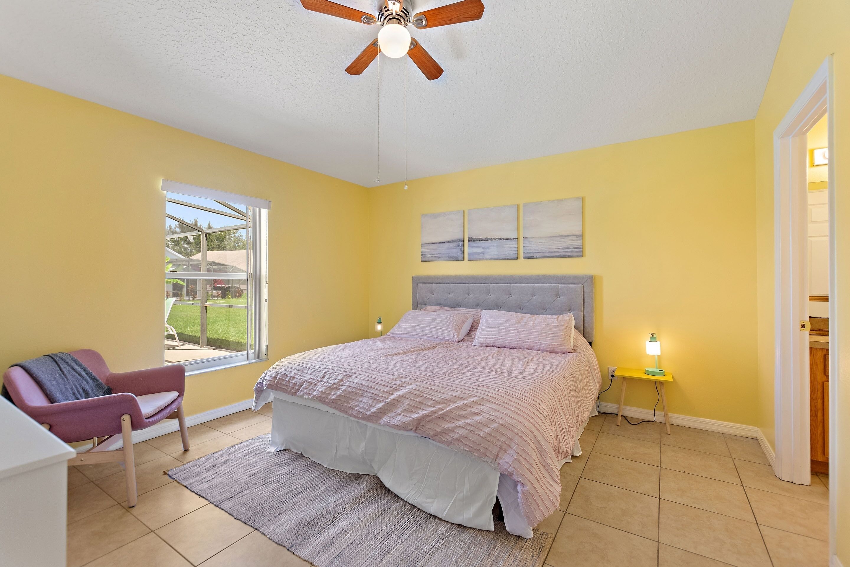 Quarto com cama, uma cadeira, mesa de cabeceira, ventoinha no teto e janela com vista para o exterior.