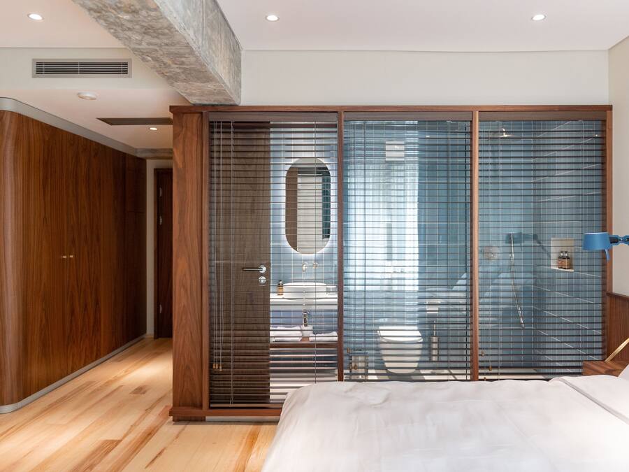 Quarto moderno com guarda-roupa de madeira, banheiro com divisória de vidro e luminária de cabeceira.