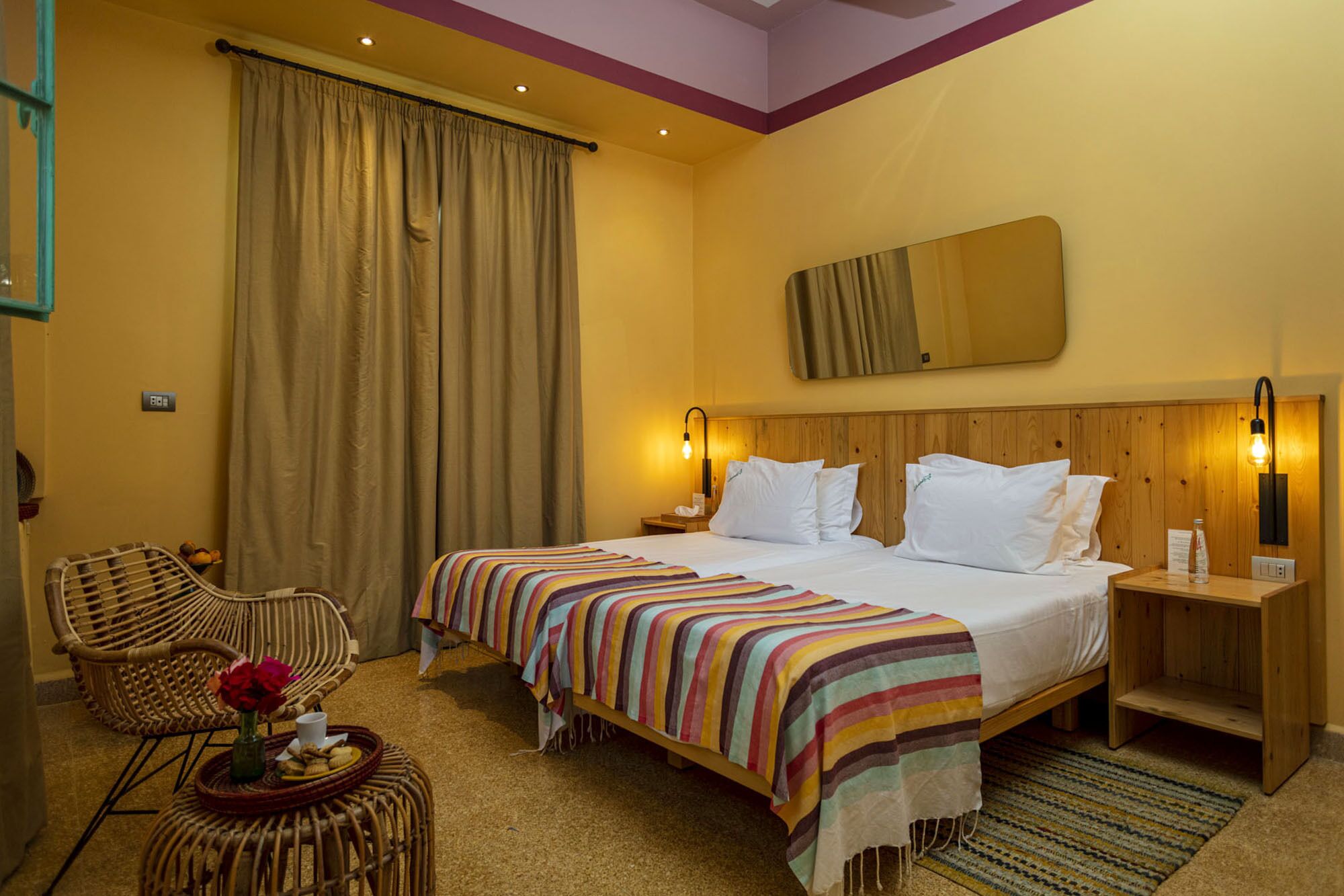 Quarto de hotel com cama, cabeceira de madeira, mesa de cabeceira, cadeira de vime, pequena mesa com um vaso e um cesto, e uma grande janela com cortinas.