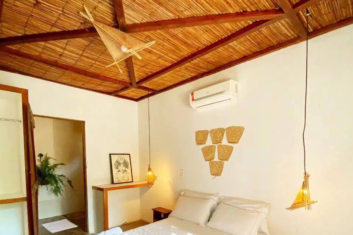 Quarto com estrutura de cama em madeira, roupa de cama branca, ventilador de teto, ar condicionado fixado na parede e uma pequena mesa de cabeceira com luminária.
