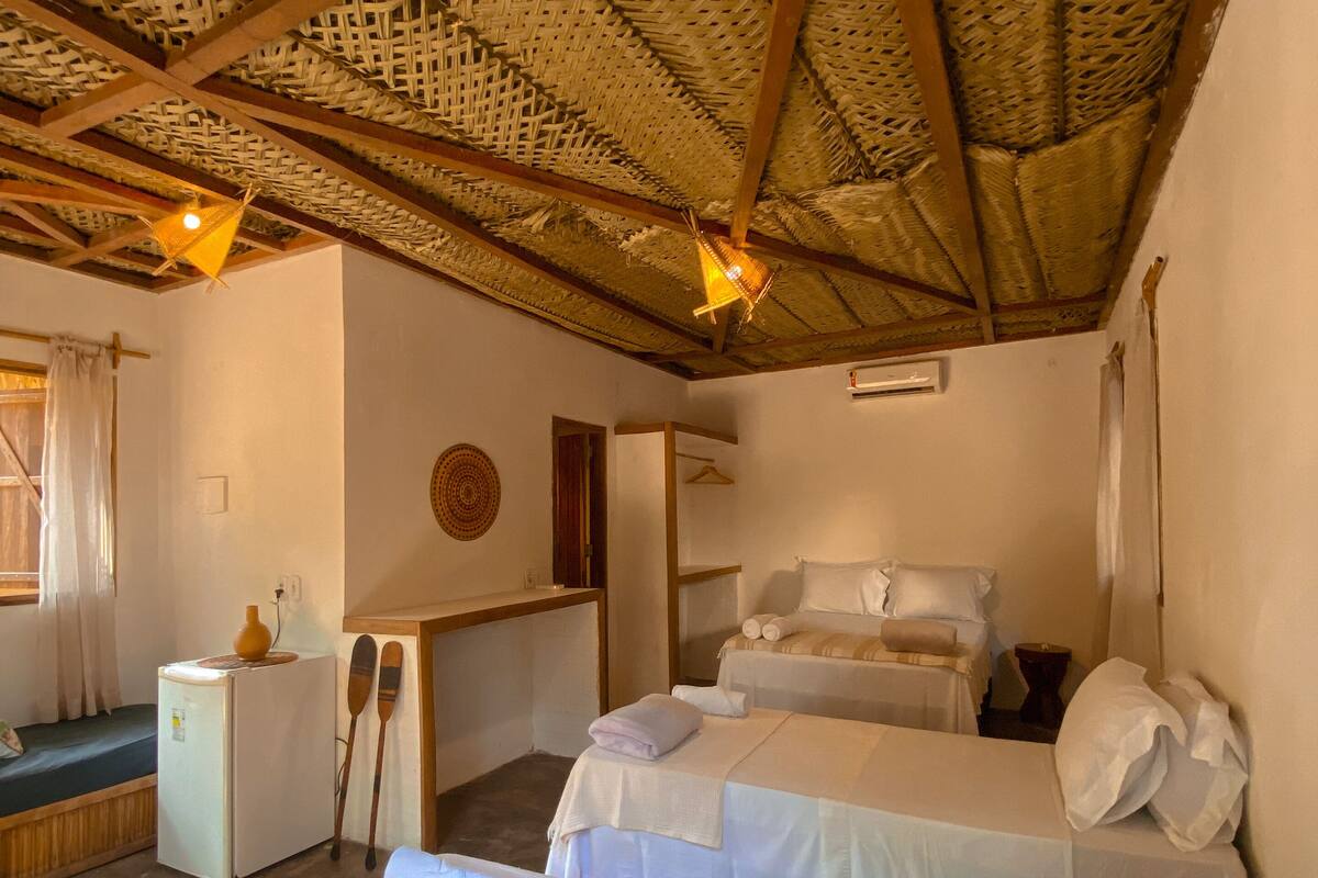 Quarto com duas camas, uma escrivaninha pequena, um refrigerador e teto de palha.