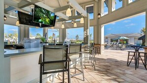Uma área de bar ao ar livre ampla, com pé-direito alto, grandes janelas e uma tela de televisão.