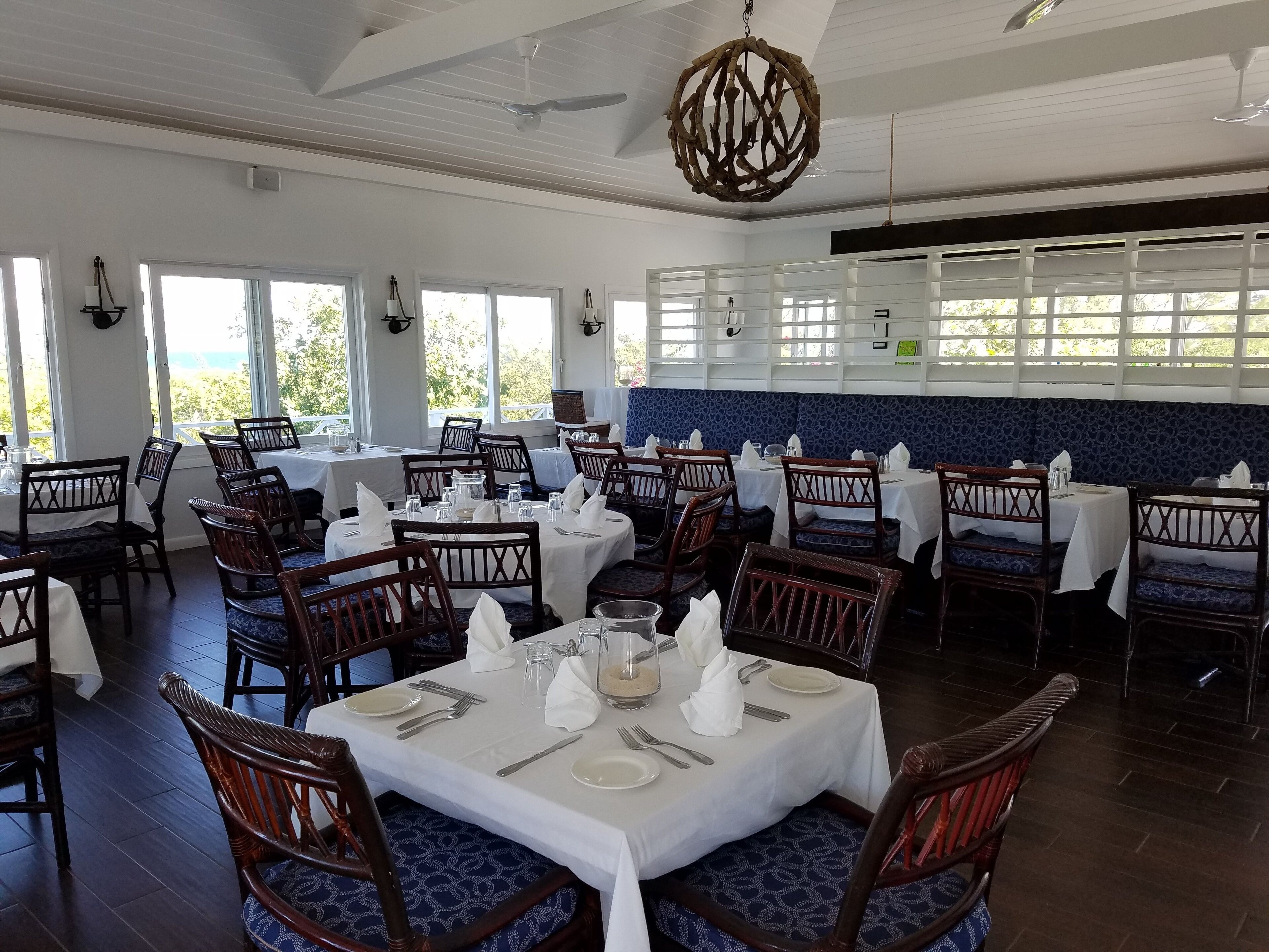 Um restaurante com várias mesas e cadeiras, janelas amplas com vista e um ventilador de teto.