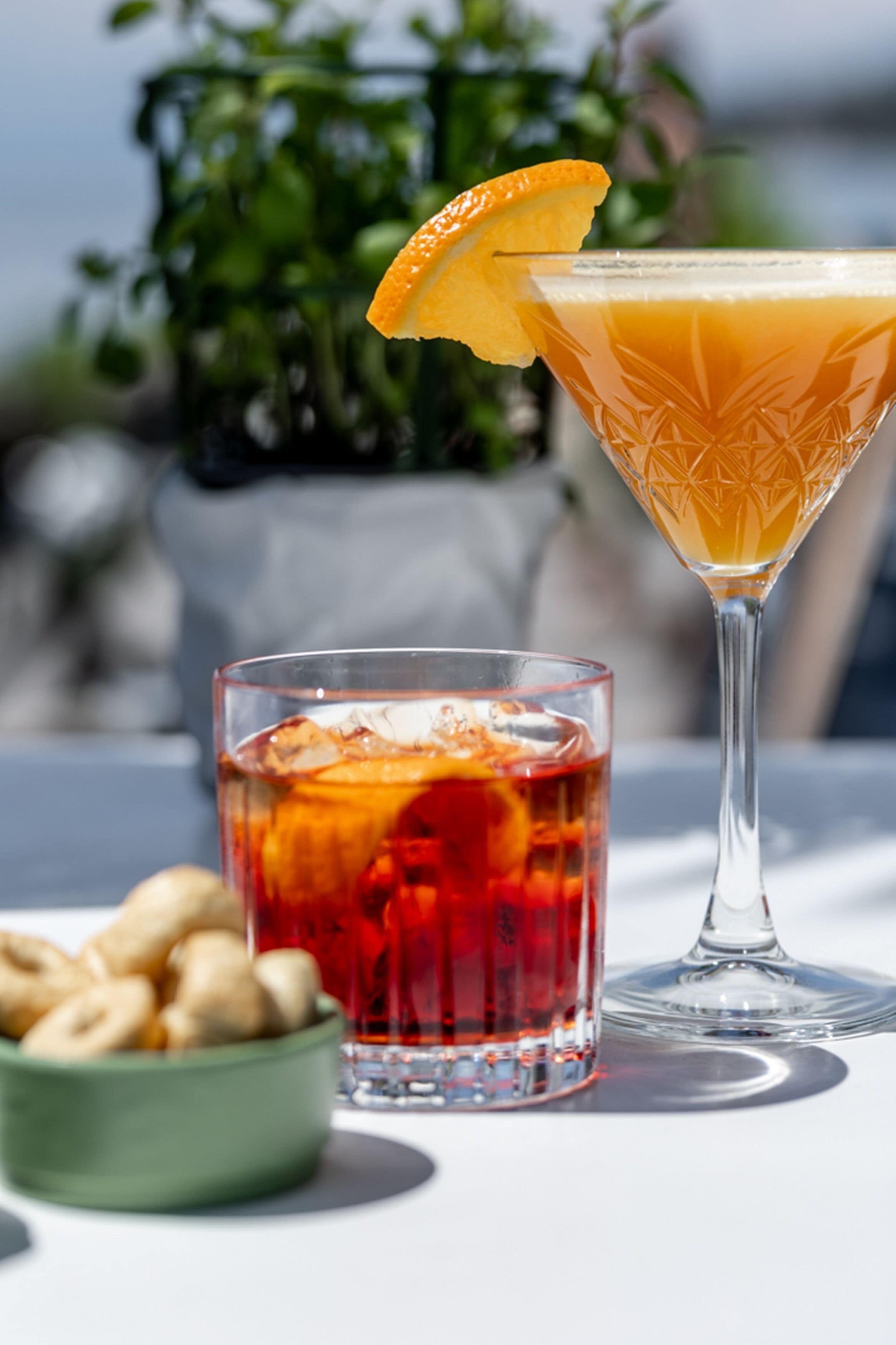 Duas bebidas, uma com cubos de gelo e outra com um pedaço de laranja, sobre uma mesa com uma tigela de petiscos.