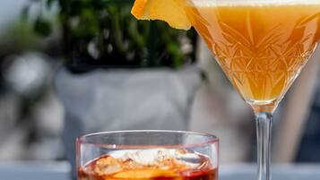 Duas bebidas, uma com cubos de gelo e outra com um pedaço de laranja, sobre uma mesa com uma tigela de petiscos.