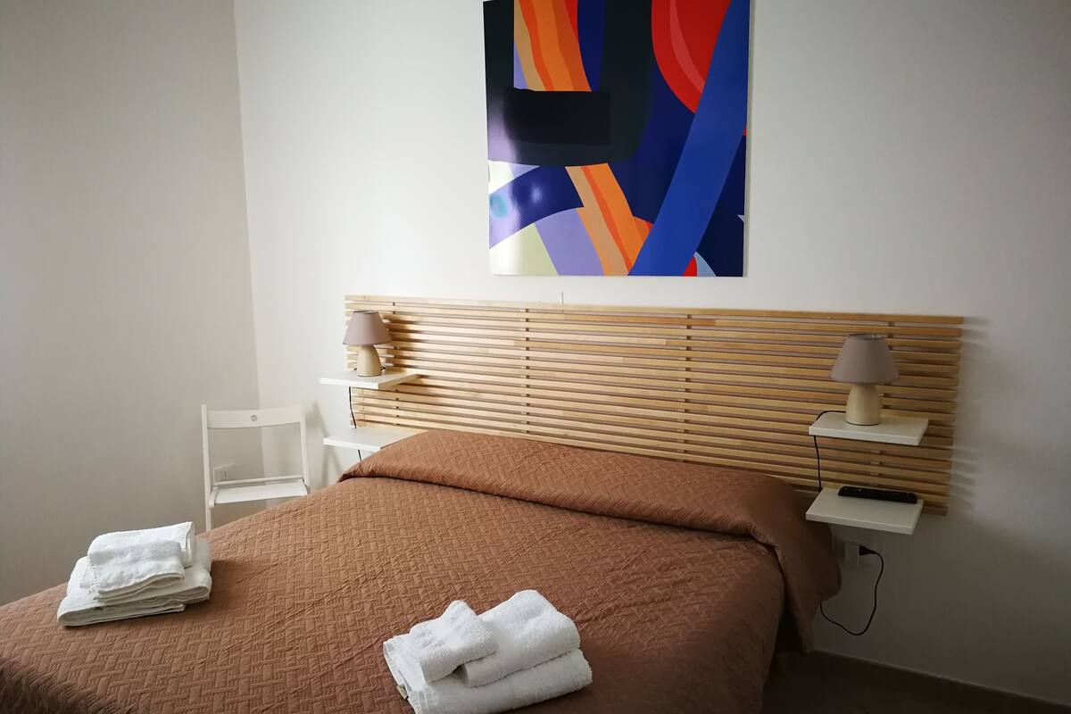 Quarto de hotel com cabeceira de madeira, uma grande pintura abstrata, cama com colcha marrom, duas mesas de cabeceira com abajures e uma toalha branca dobrada na cama.