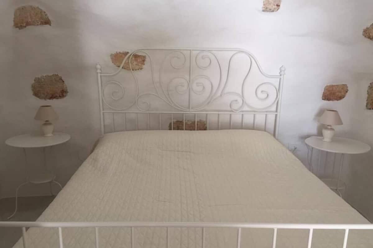 Uma cama com estrutura de metal branca e colcha bege com textura. Existem duas mesinhas de cabeceira brancas com abajures de cada lado da cama.