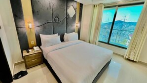 Quarto de hotel moderno com uma cama grande, uma mesa de cabeceira com telefone e vista para a cidade através de amplas janelas.
