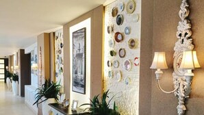 Corredor de hotel com um longo balcão de recepção, plantas em vasos, quadros e uma parede decorada com pratos e um abajur.