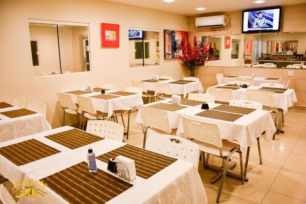 Uma área de refeições de hotel com várias mesas preparadas para hóspedes, uma televisão na parede e um espelho refletindo parte do espaço.