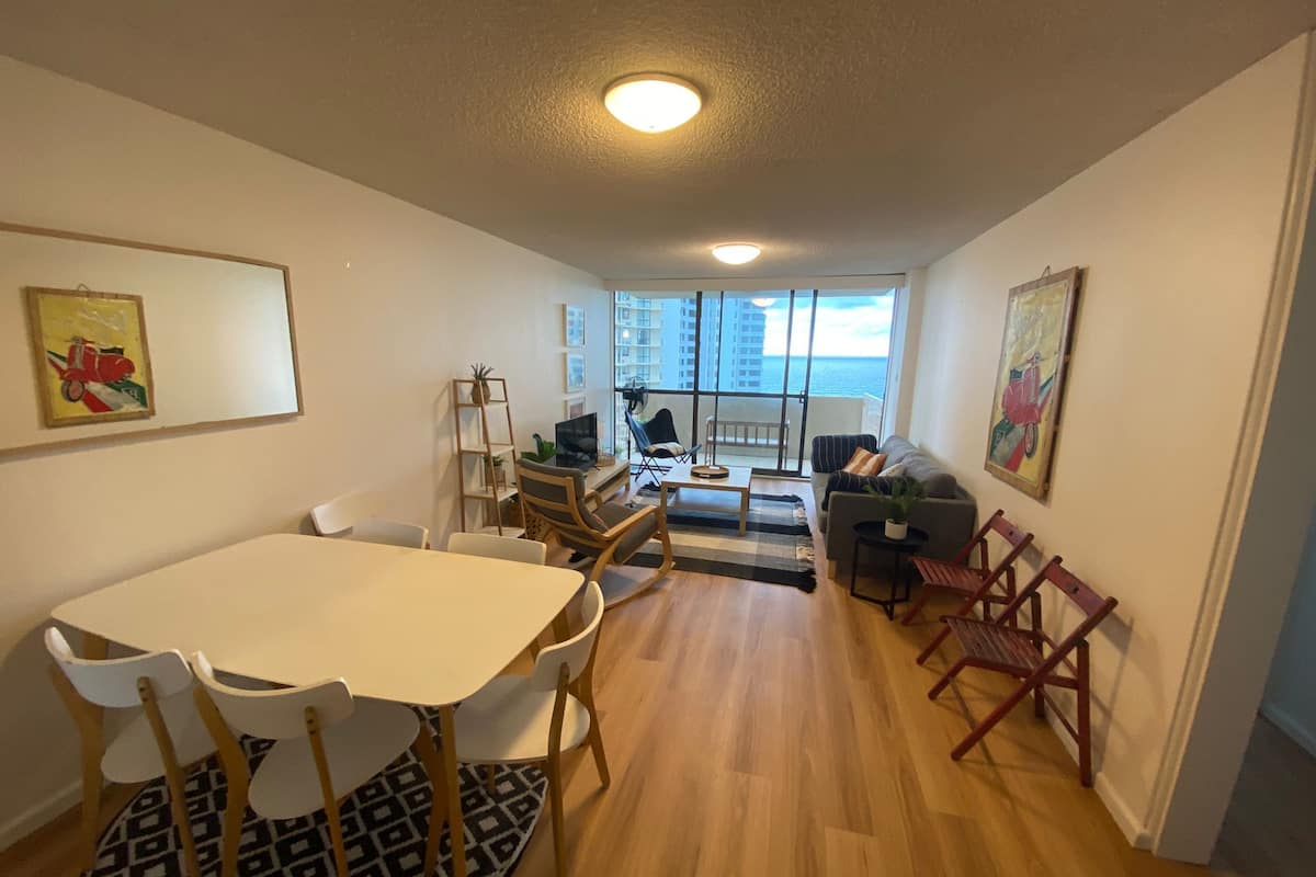 Sala de estar moderna com área de jantar, sofá, mesa pequena e vista para o oceano através de grandes janelas.