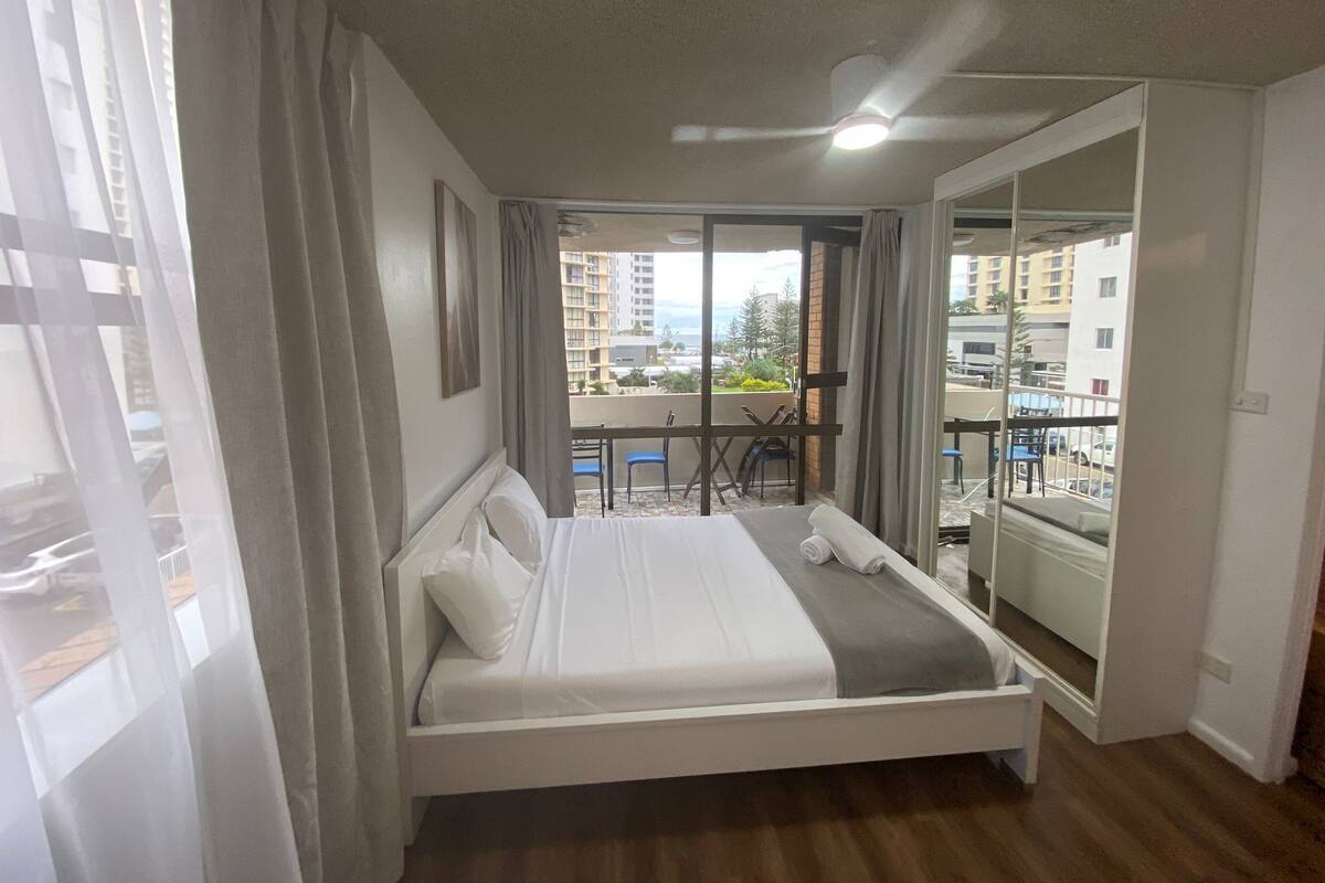 Quarto com cama, ventilador de teto, janela com cortinas e vista para a área da varanda.