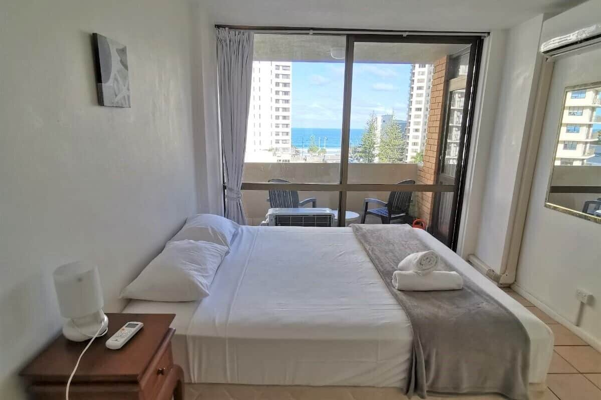 Quarto de hotel com cama, mesa de cabeceira com abajur, janela com vista para edifícios e o mar, e uma luminária no teto.