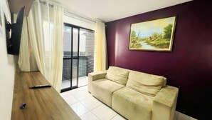 Uma sala de estar com um sofá bege, um quadro na parede e uma porta de vidro deslizante que dá para uma varanda.