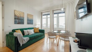 Uma sala de estar com um sofá verde, uma mesa de jantar de vidro com cadeiras, uma televisão e obras de arte na parede.