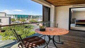 Uma varanda com uma mesa redonda e cadeiras, um vaso de planta e vista para um edifício moderno e vegetação.