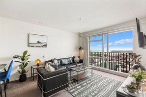 Sala de estar moderna com sofá de couro preto, mesa de centro e vista da cidade através de grandes janelas.
