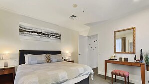 Quarto moderno com uma cama grande, mesas de cabeceira, escrivaninha com cadeira, espelho e um objeto decorativo na parede.