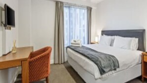 Quarto de hotel com cama, escrivaninha, cadeira, televisão e janela com cortinas.