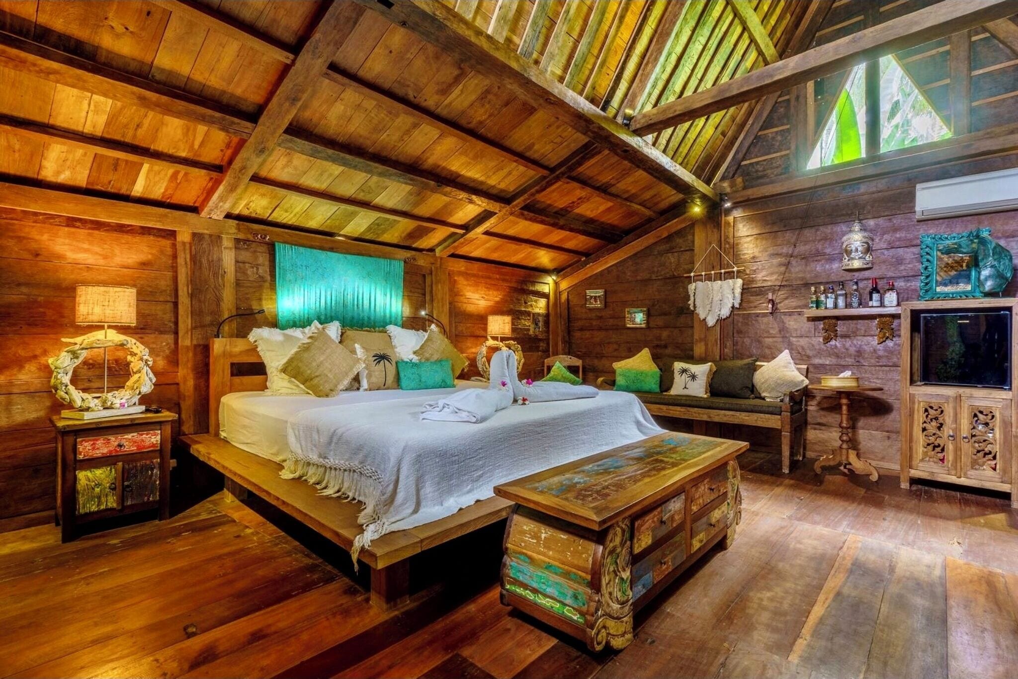Um quarto de madeira com uma cama, um banco de madeira, uma mesinha e um armário.