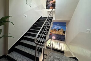 Uma escadaria com corrimão de metal, um interruptor de luz fixado na parede e um cartaz de um edifício com água e céu.
