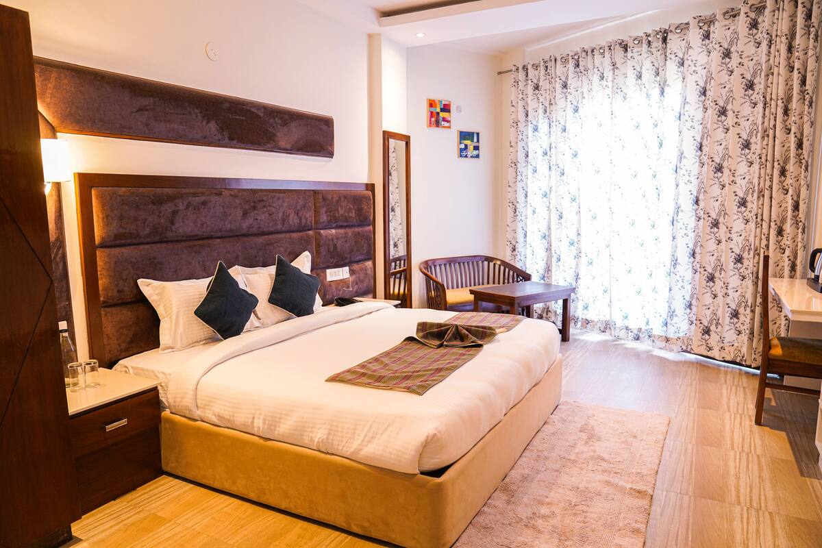 Quarto de hotel com uma cama grande, cabeceira de madeira, mesa de cabeceira com abajur, uma cadeira e uma pequena mesa com espelho.