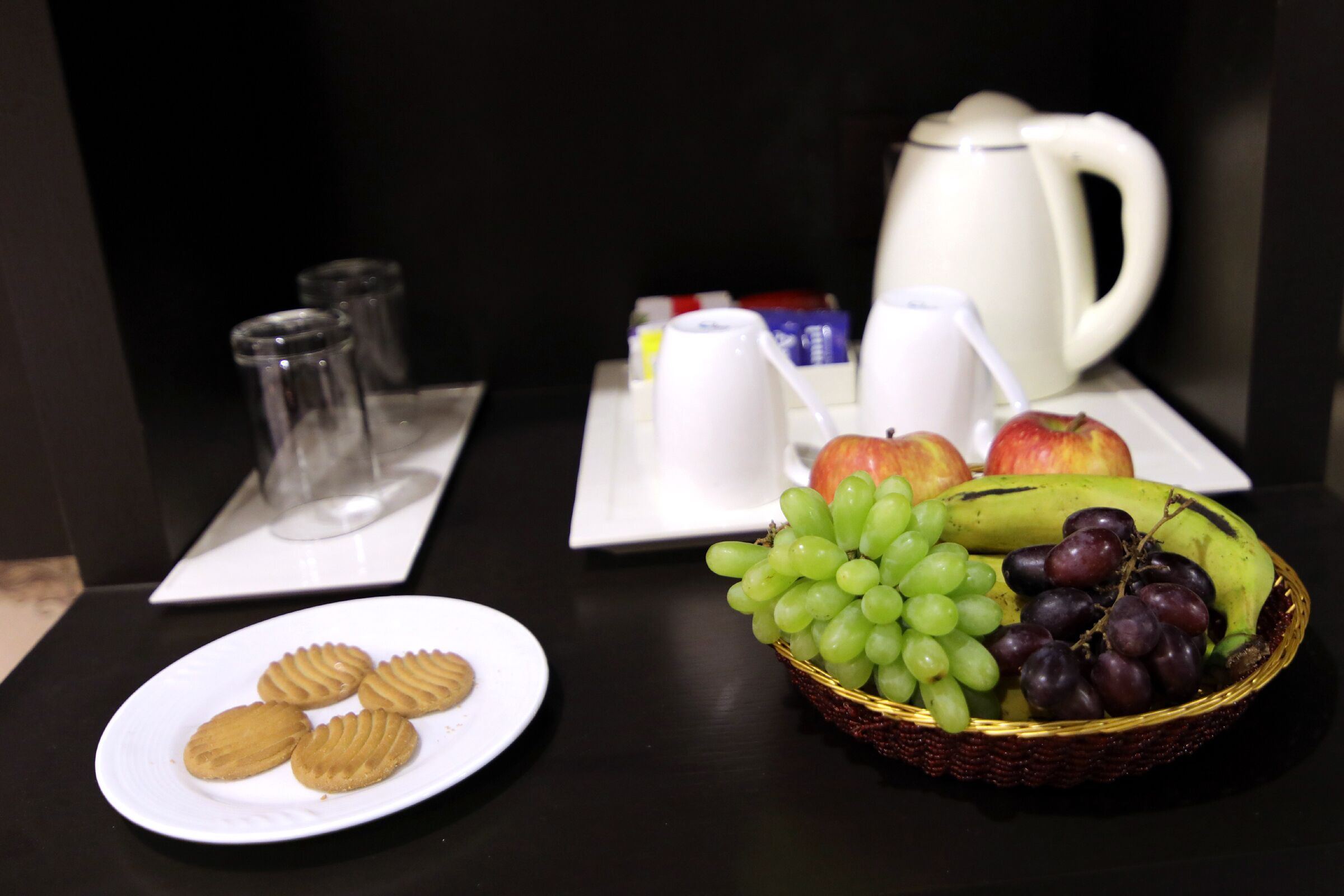 Mesa preta com arrumação de café da manhã, incluindo um prato de biscoitos, cesta de frutas, cafeteira branca e jarra de vidro.