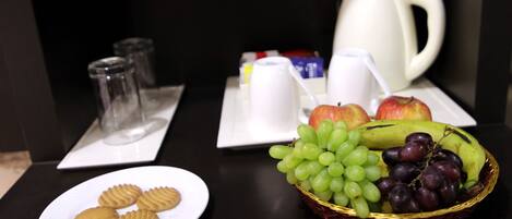 Mesa preta com arrumação de café da manhã, incluindo um prato de biscoitos, cesta de frutas, cafeteira branca e jarra de vidro.