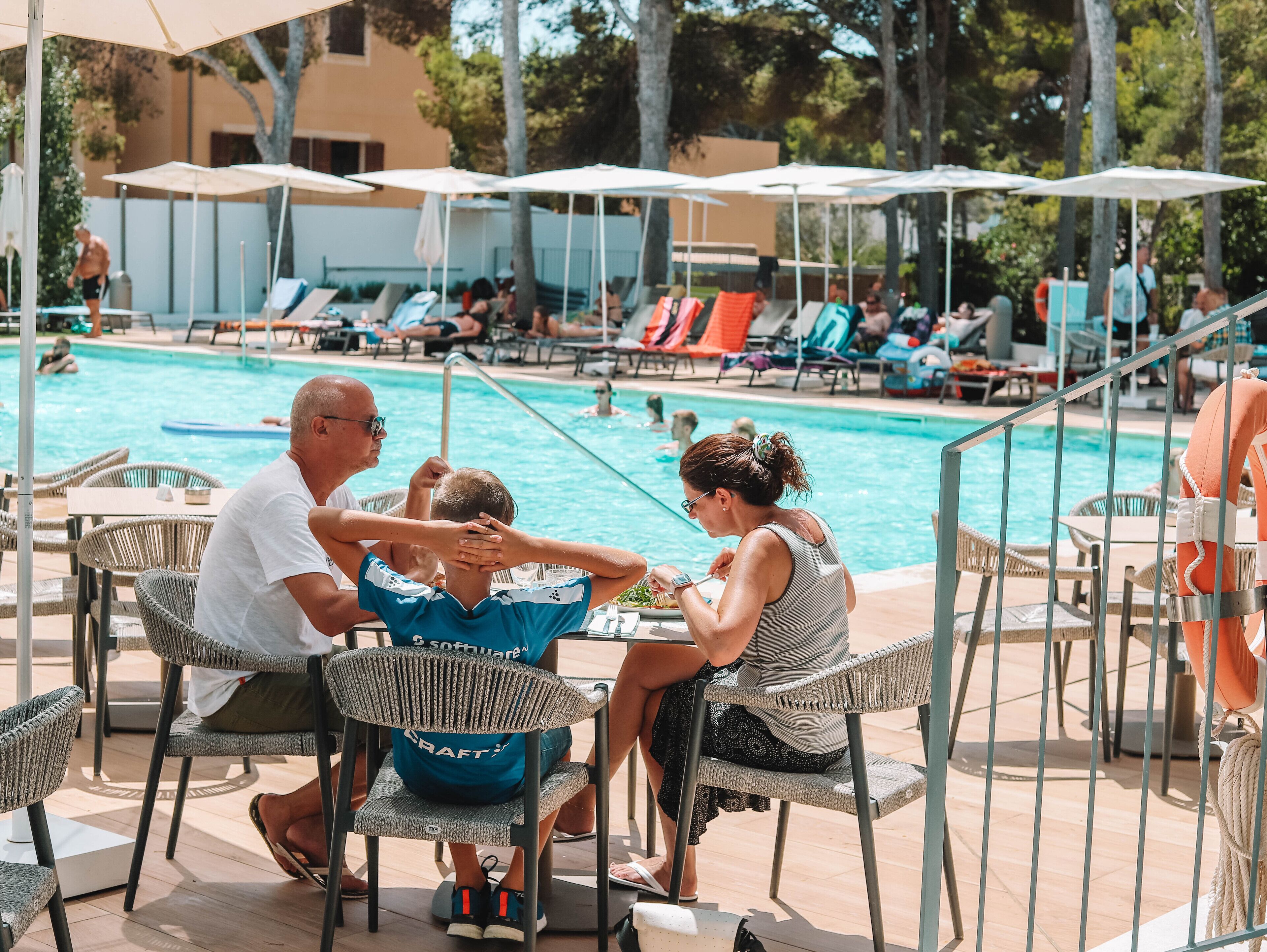 Uma área à beira da piscina com espreguiçadeiras e guarda-sóis, uma mesa de jantar com cadeiras e a piscina ao fundo.