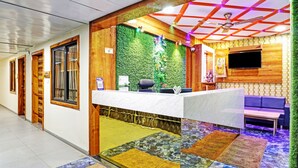 Uma área de recepção com balcão de mármore, parede verde com plantas, ventilador de teto e televisão fixada na parede.