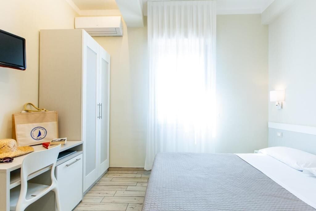 Quarto de hotel com cama, escrivaninha com cadeira, guarda-roupa, televisão na parede e janela com cortinas.