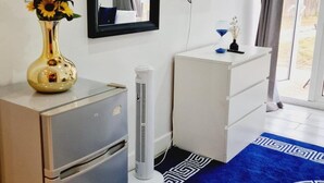 Quarto compacto com geladeira, cômoda branca, ventilador e tapete azul estampado.