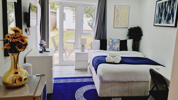 Quarto de hotel com cama, escrivaninha com um vaso de girassóis, cadeira, mesinha de cabeceira com abajur e vista para o exterior.