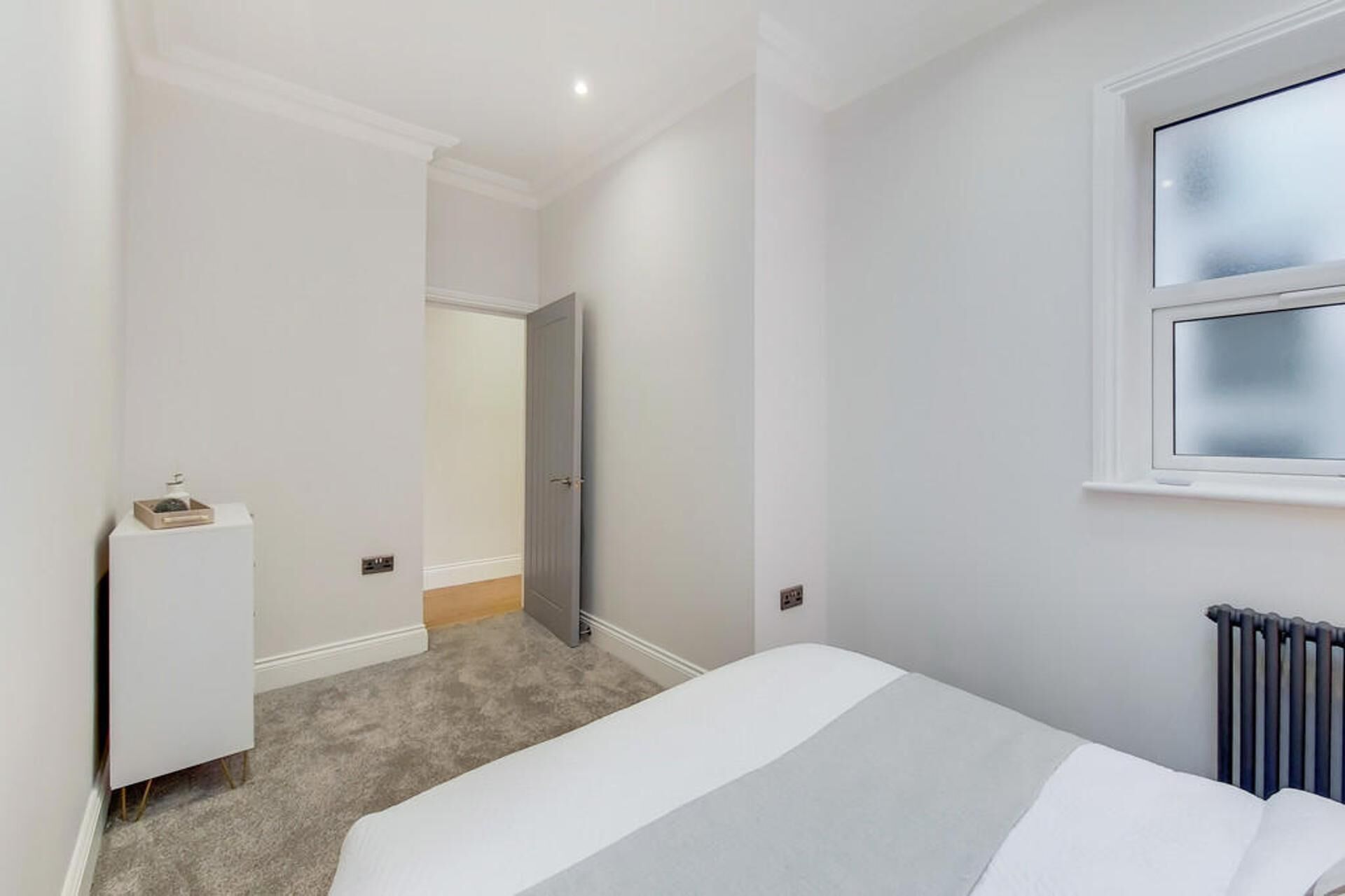 Quarto com cama, radiador, janela e porta que dá para outro cômodo.
