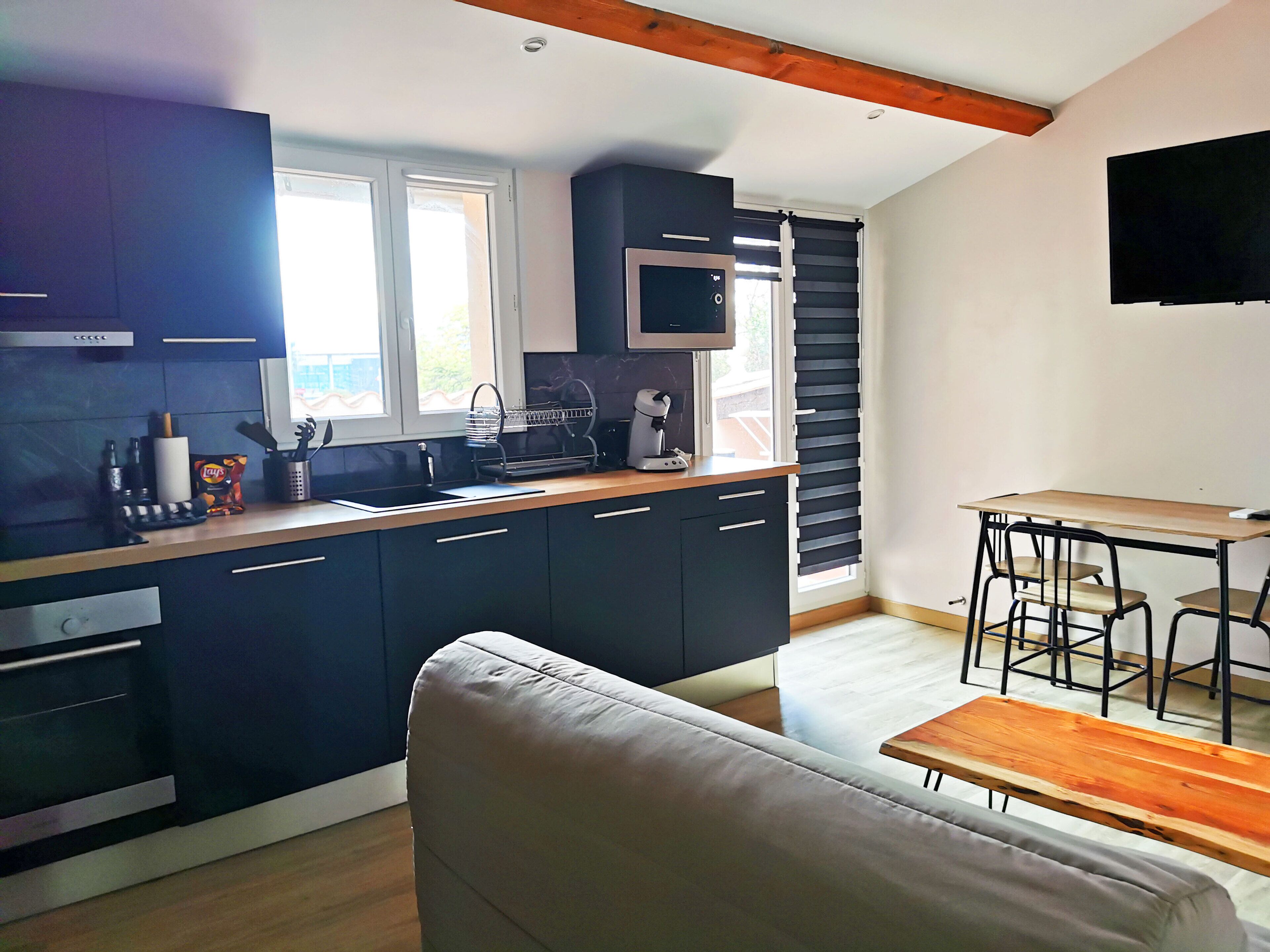 Cozinha moderna com armários escuros, bancada de madeira e micro-ondas embutido. Há uma área de refeições com mesa e cadeiras de madeira, e uma janela grande que oferece luz natural.