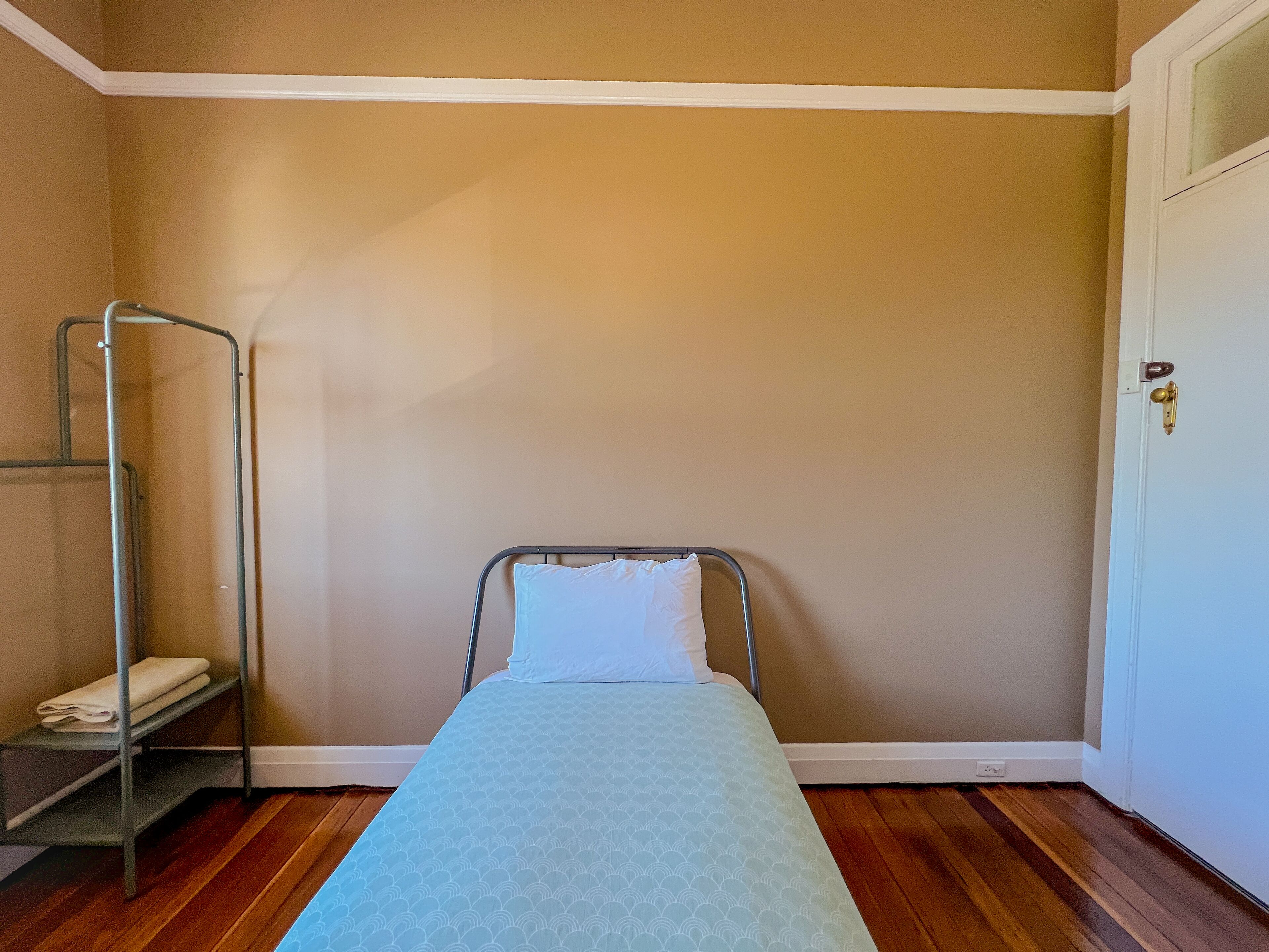 Um quarto pequeno com uma cama de solteiro e um beliche à esquerda, uma porta branca à direita e soalho de madeira.