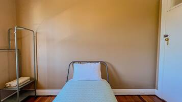 Um quarto pequeno com uma cama de solteiro e um beliche à esquerda, uma porta branca à direita e soalho de madeira.
