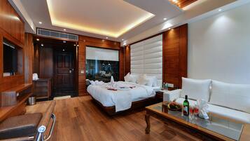 Quarto de hotel moderno com piso de madeira, uma cama grande com roupa de cama branca, um sofá branco, uma mesa de centro de vidro e uma mesinha de cabeceira de madeira.