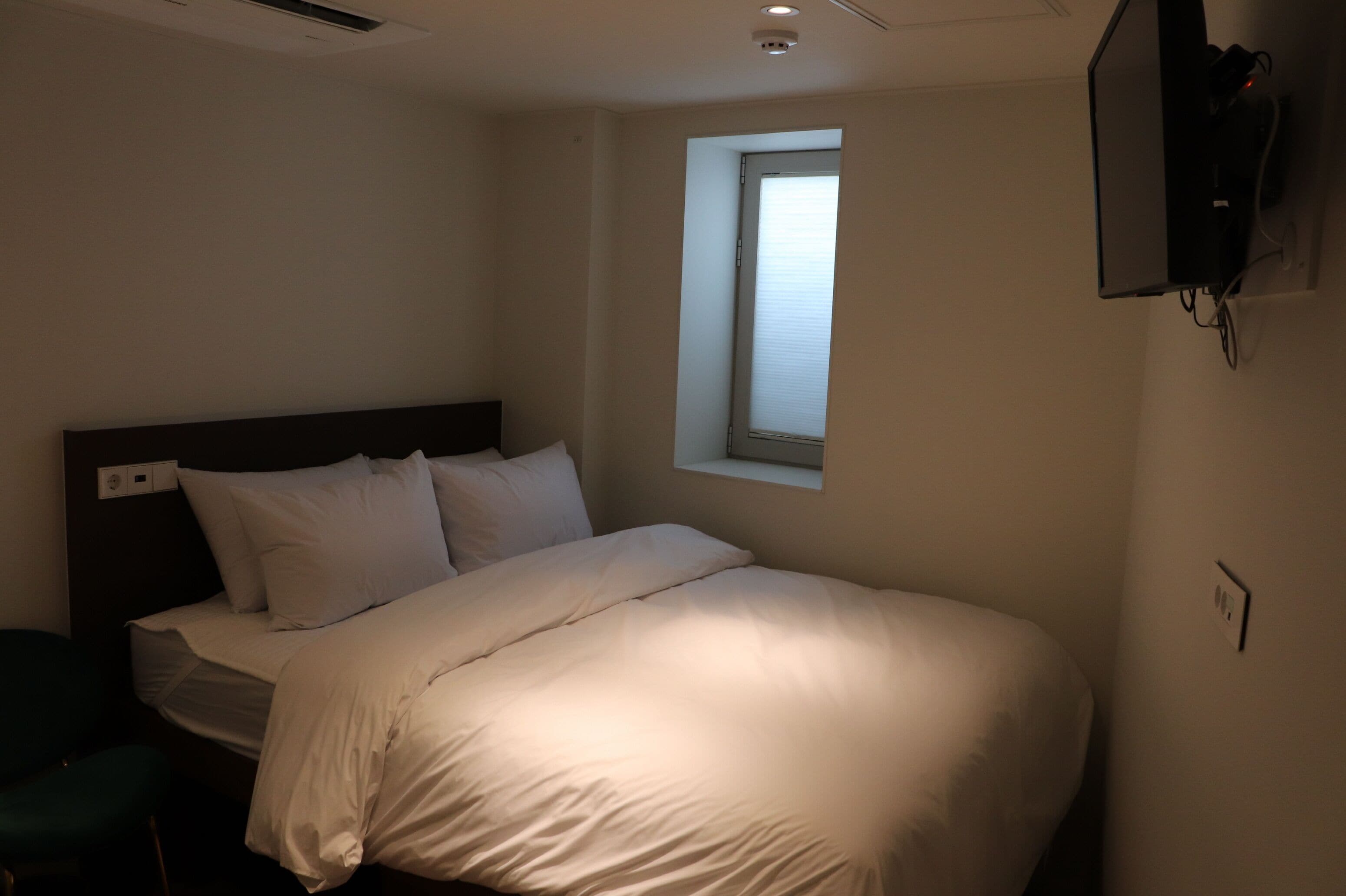 Quarto de hotel com cama, janela e televisão presa na parede.