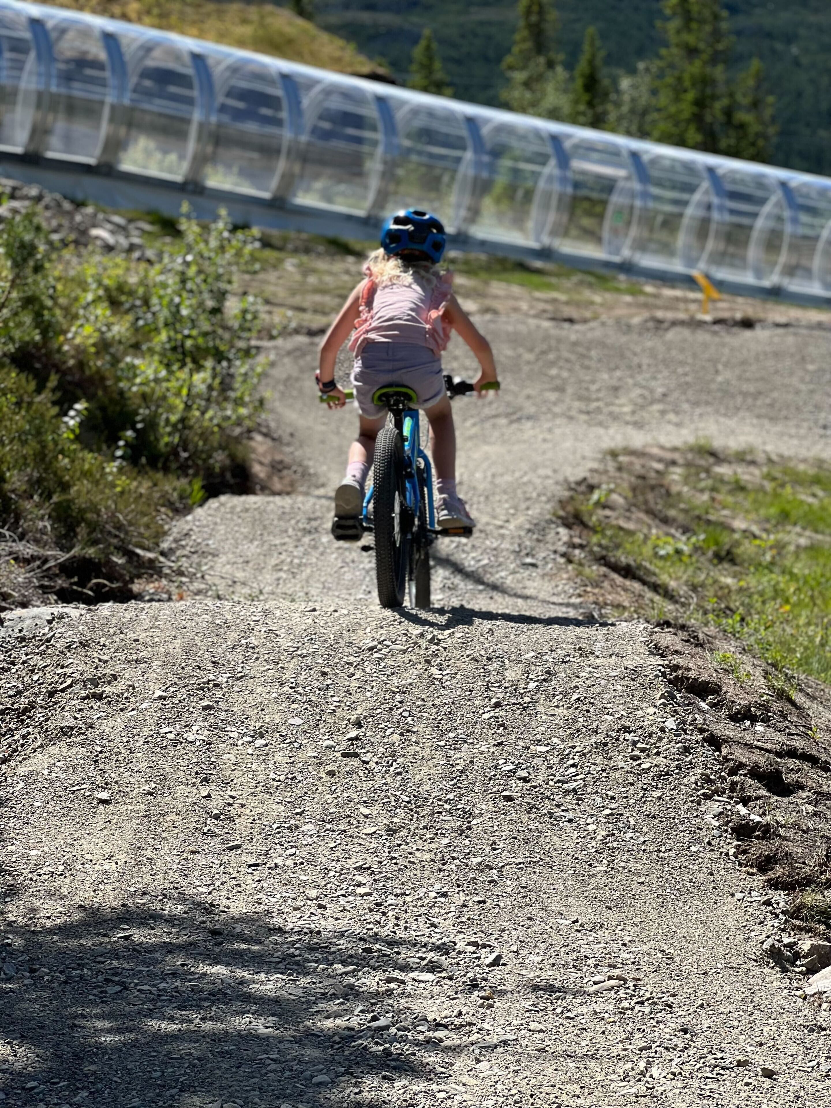 Uma criança anda de bicicleta por um caminho de cascalho, com uma cerca ao fundo.