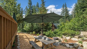 Um deck de madeira com mesa para piquenique e guarda-sol, rodeado por árvores e um cercado de madeira.