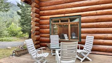 Uma cabana de toras com varanda, mobiliário para área externa e janela com vista para uma área arborizada.