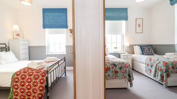 Quarto de hotel com duas camas, uma janela com cortina azul, mesa de cabeceira com abajur e relógio fixado na parede.