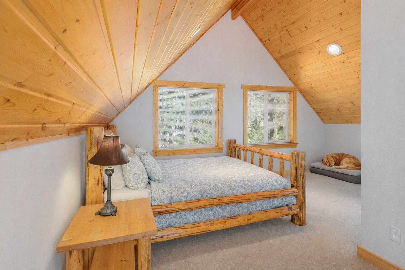 Um quarto de sótão acolhedor com uma cama de madeira, duas janelas com persianas, uma mesa de cabeceira com abajur e um cão descansando sobre um travesseiro.