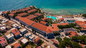 Vista aérea de um resort à beira-mar, com piscina, praia e uma fileira de vilas com telhados vermelhos.