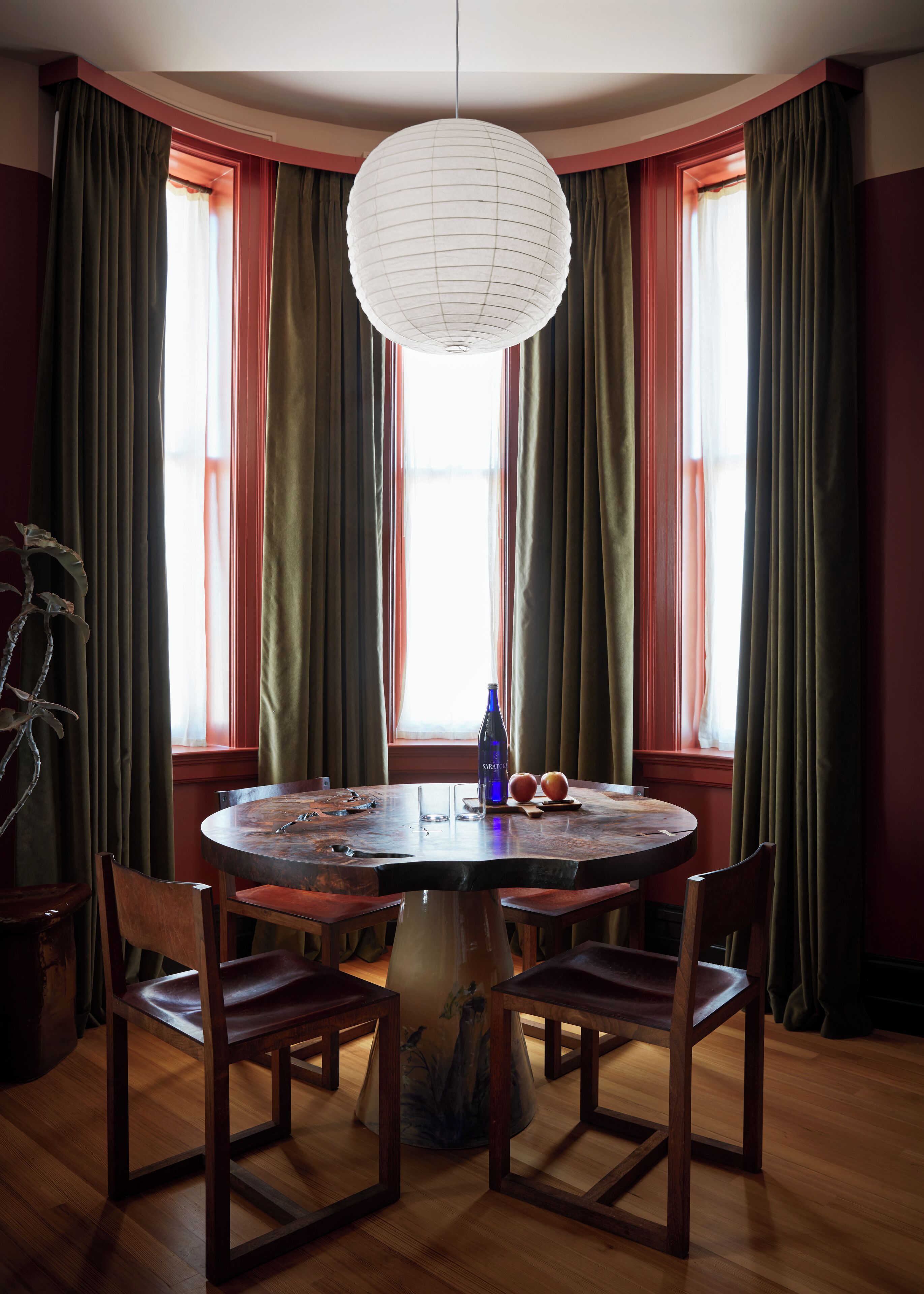 Uma sala de jantar com mesa redonda, quatro cadeiras, luminária pendente e janela com cortinas.