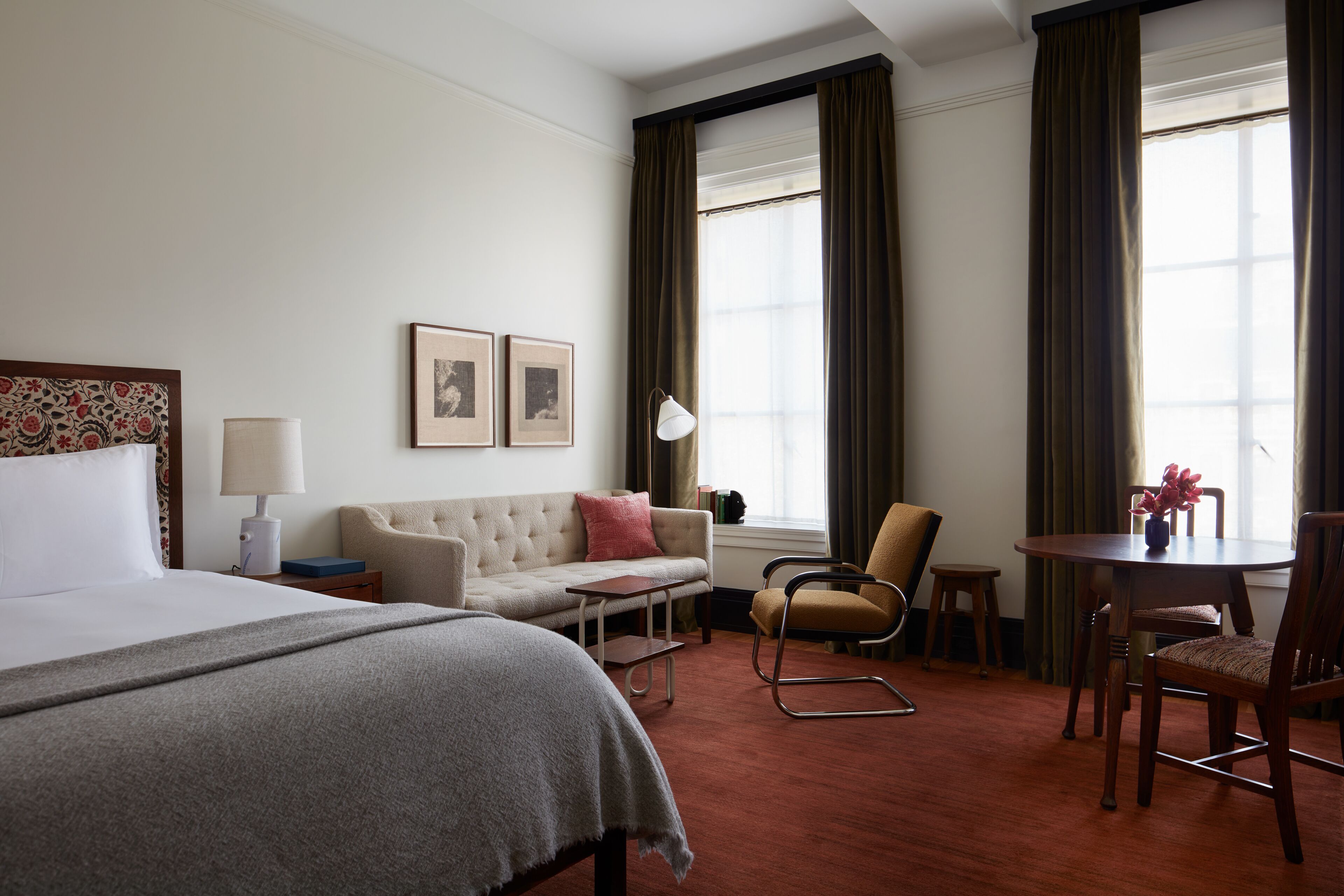Quarto de hotel com cama, sofá, mesa de jantar e uma cadeira. Há uma janela com cortinas, um abajur e quadros na parede.