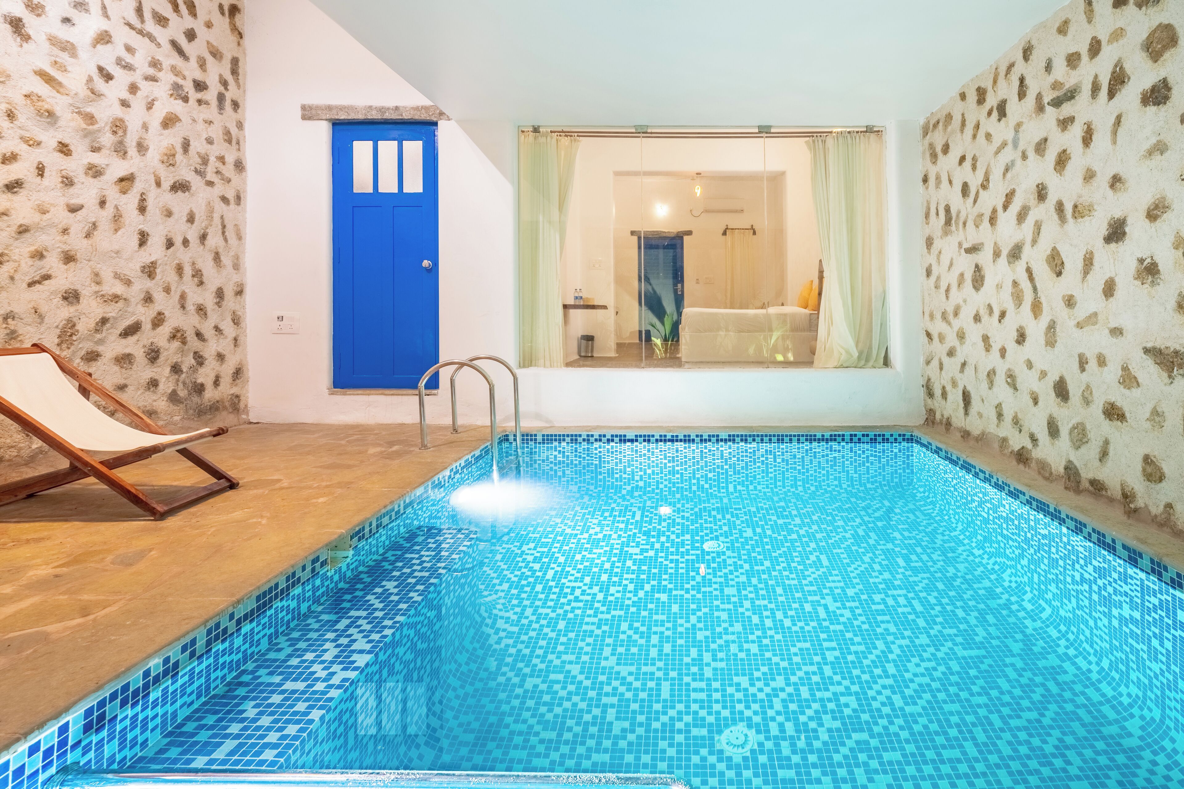 Piscina com chão ladrilhado, porta azul e vista para um quarto com cama e cortinas.