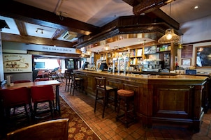 Uma área de bar com banquetas de madeira, piso ladrilhado e diversas garrafas e copos em prateleiras.