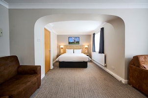 Quarto de hotel com uma cama grande, duas mesas de cabeceira com luminárias, cabeceira, janela com cortinas e piso coberto por carpete.